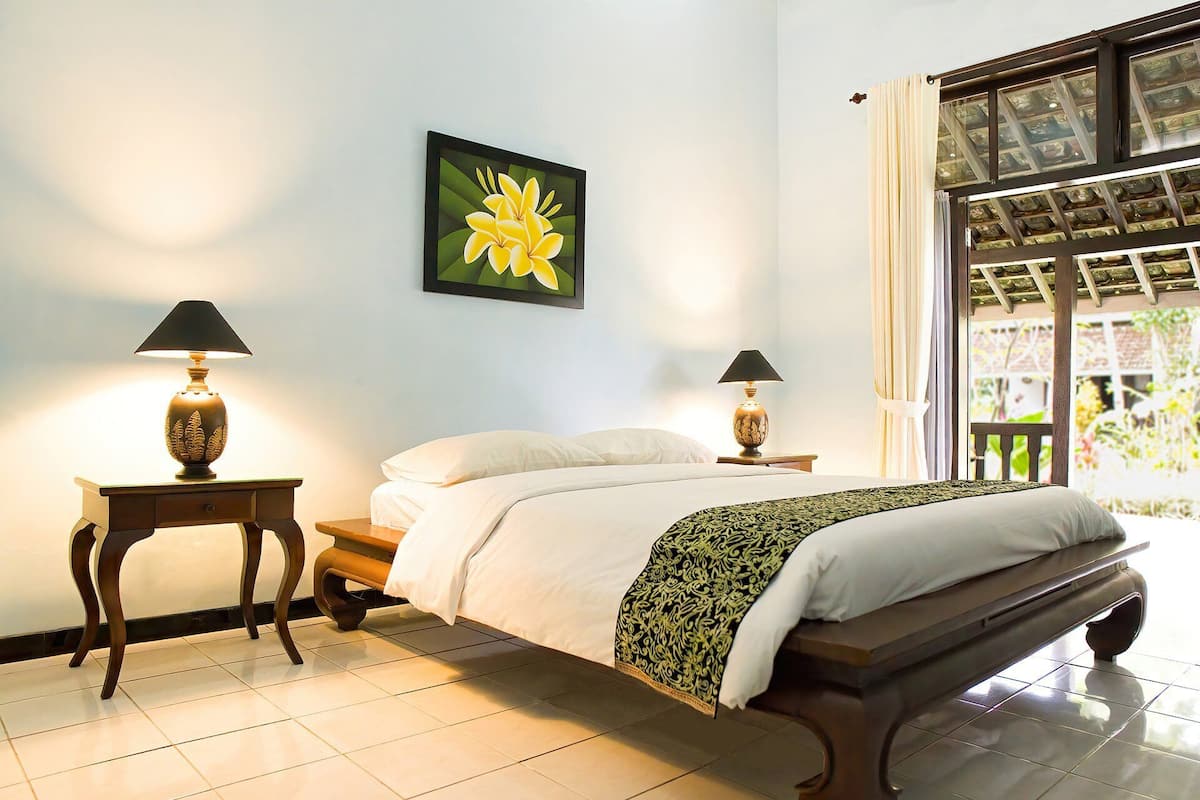 Quarto com cama, duas mesas de cabeceira com luminárias, uma janela grande com cortinas e um quadro de flor na parede.