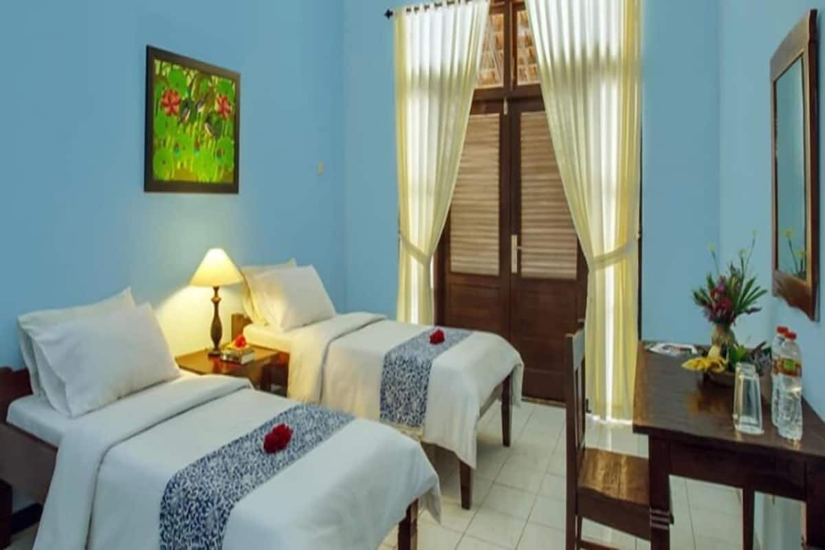 Quarto de hotel com duas camas, uma mesinha de cabeceira com abajur, uma porta de madeira, uma mesa com um vaso e garrafas, e um quadro na parede.