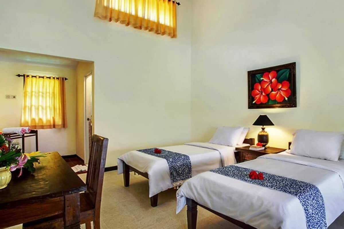 Quarto de hotel com duas camas de solteiro, uma mesa de madeira, uma cadeira, um quadro com flores vermelhas e um abajur.
