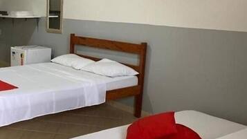 Quarto com duas camas de madeira, roupa de cama branca e um travesseiro vermelho em uma das camas. Há um pequeno armário branco e um espelho na parede.