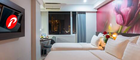 Quarto de hotel com duas camas, uma escrivaninha, uma cadeira, televisão e uma grande janela com vista para a cidade.