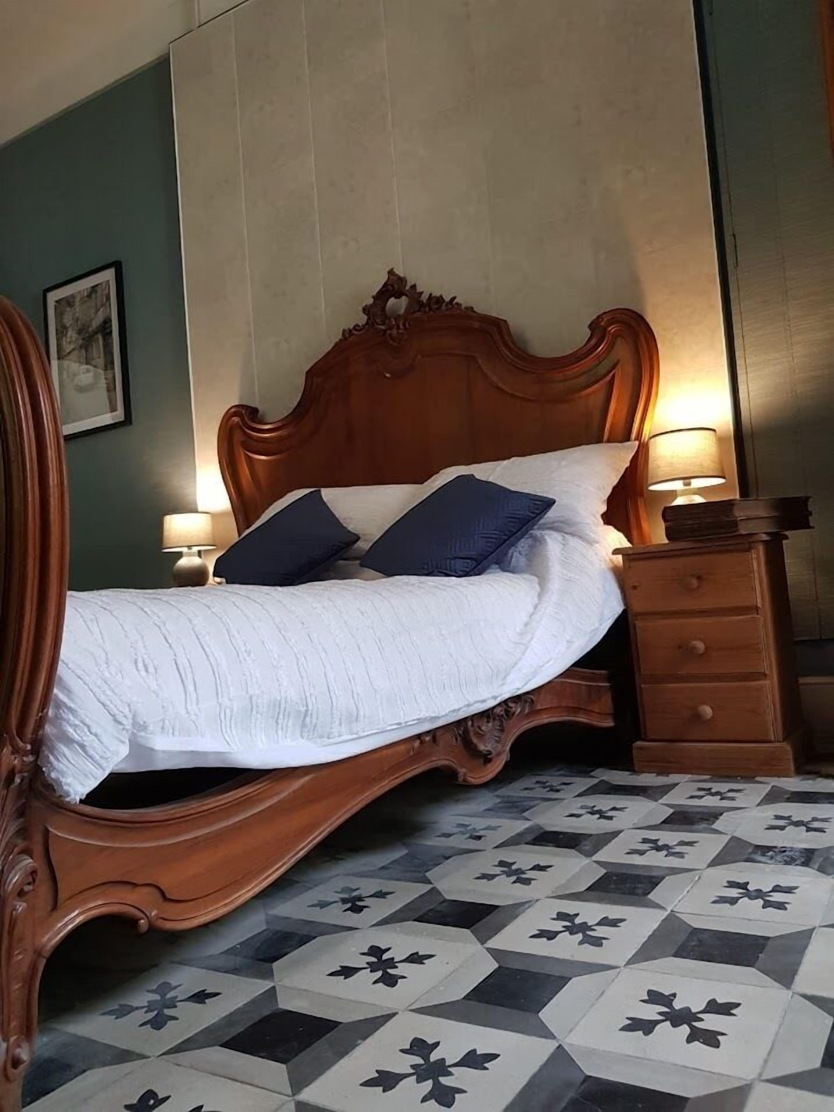 Quarto com cama de madeira, roupa de cama branca, mesa de cabeceira com abajur e piso estampado.