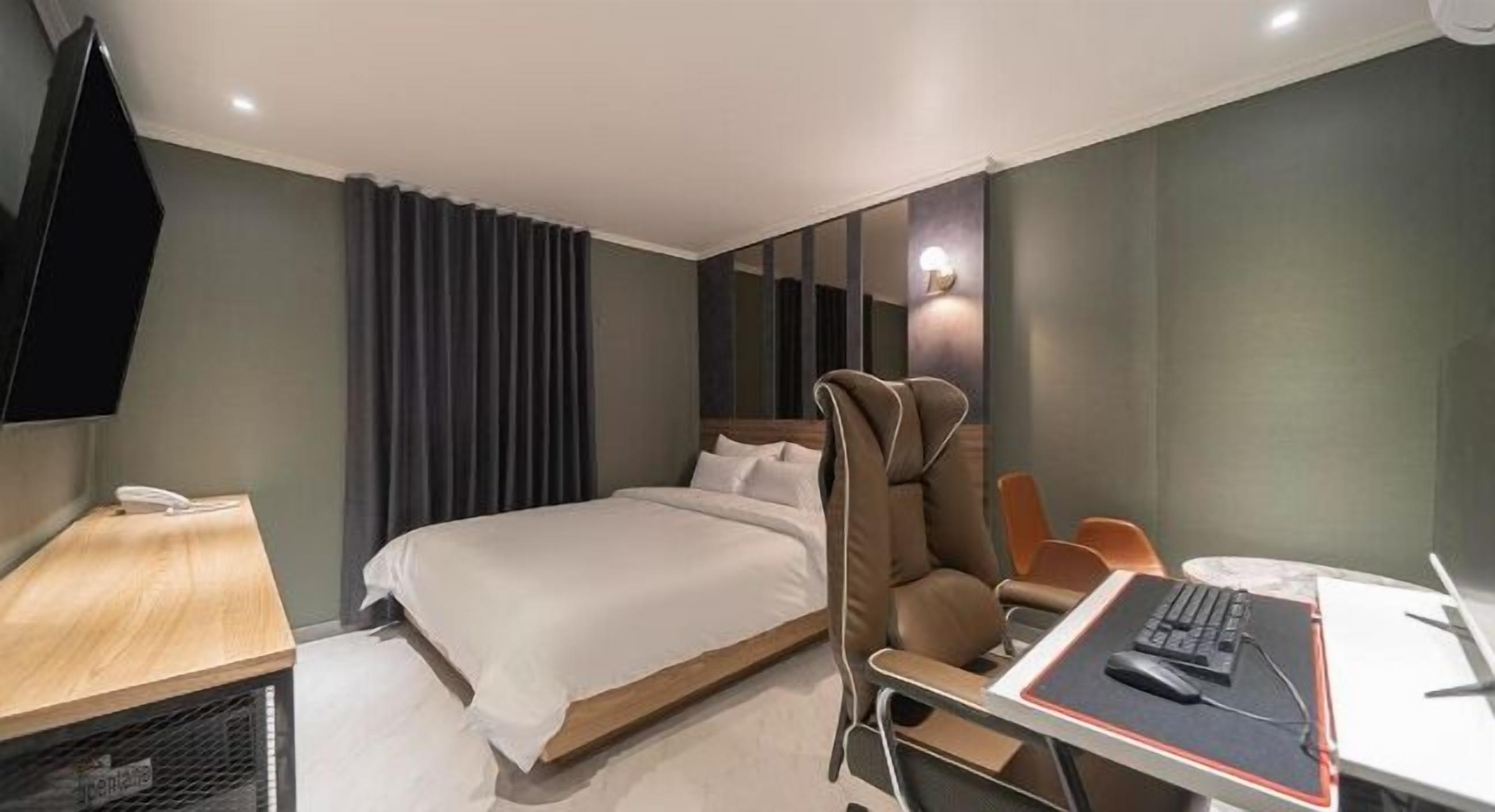 Quarto de hotel moderno com cama, secretária, computador, cadeira e televisão.