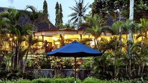 Um resort com duas espreguiçadeiras sob um guarda-sol azul, rodeado por vegetação exuberante e palmeiras, com um edifício ao fundo.