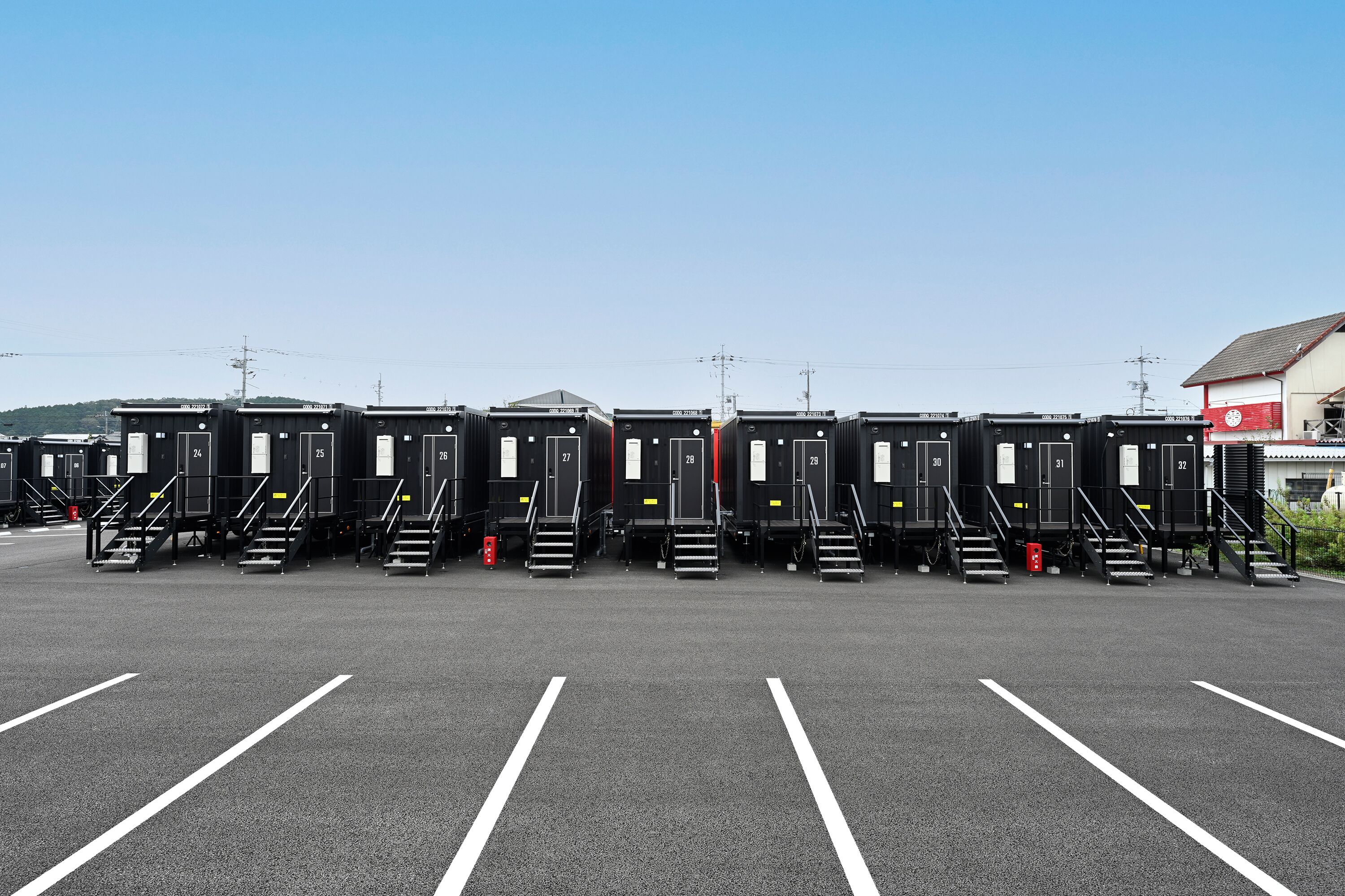 Uma fileira de sanitários portáteis pretos com números nas portas, dispostos num parque de estacionamento com linhas brancas no chão.