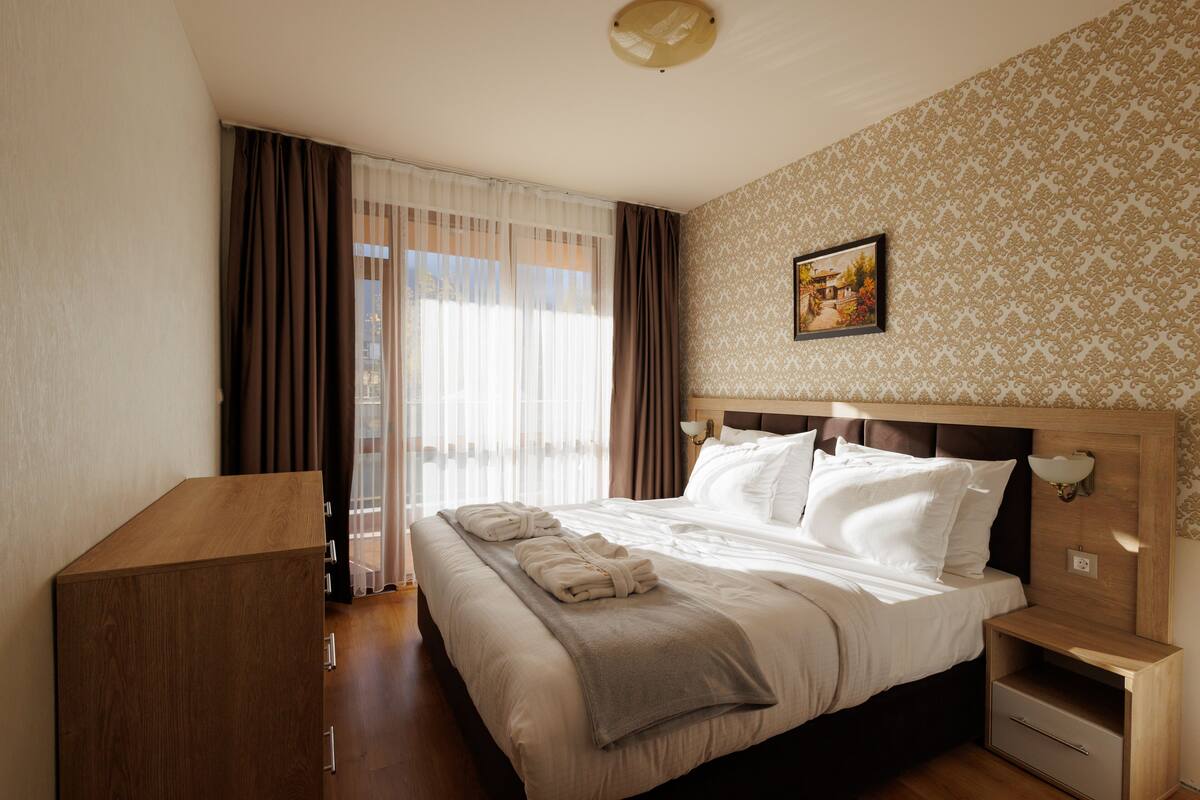Quarto de hotel com uma cama, uma escrivaninha de madeira, uma mesa de cabeceira e uma luminária fixada na parede.