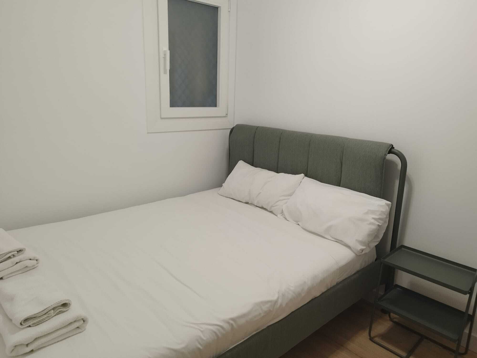 Uma cama de solteiro com cabeceira cinza, roupa de cama branca e uma pequena mesa de cabeceira com prateleira.
