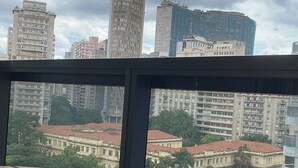 Vista de varanda com mesa e cadeiras, mostrando um horizonte urbano com diversos edifícios, incluindo um arranha-céu e um edifício histórico com telhado vermelho.