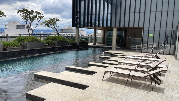 Área de piscina no terraço com espreguiçadeiras, um elemento aquático e um edifício moderno com grandes janelas.