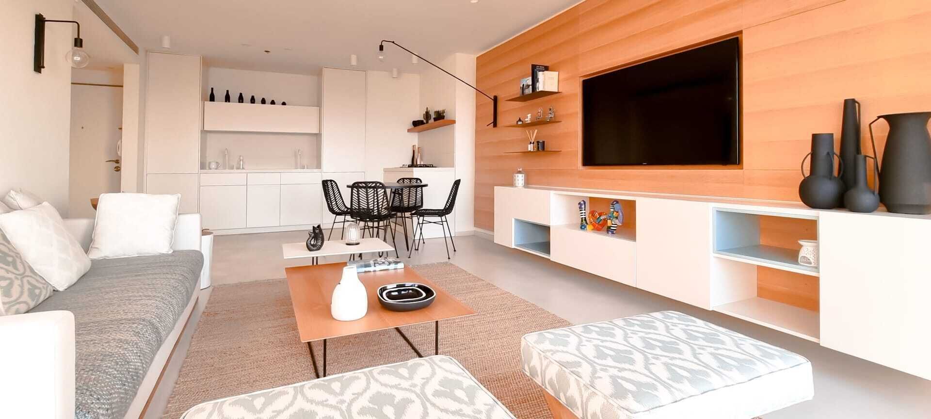 Uma sala de estar moderna com um sofá, uma mesa de centro e uma televisão fixada numa parede de madeira. Há uma área de jantar com cadeiras e uma mesa ao fundo.