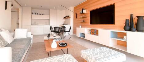 Uma sala de estar moderna com um sofá, uma mesa de centro e uma televisão fixada numa parede de madeira. Há uma área de jantar com cadeiras e uma mesa ao fundo.