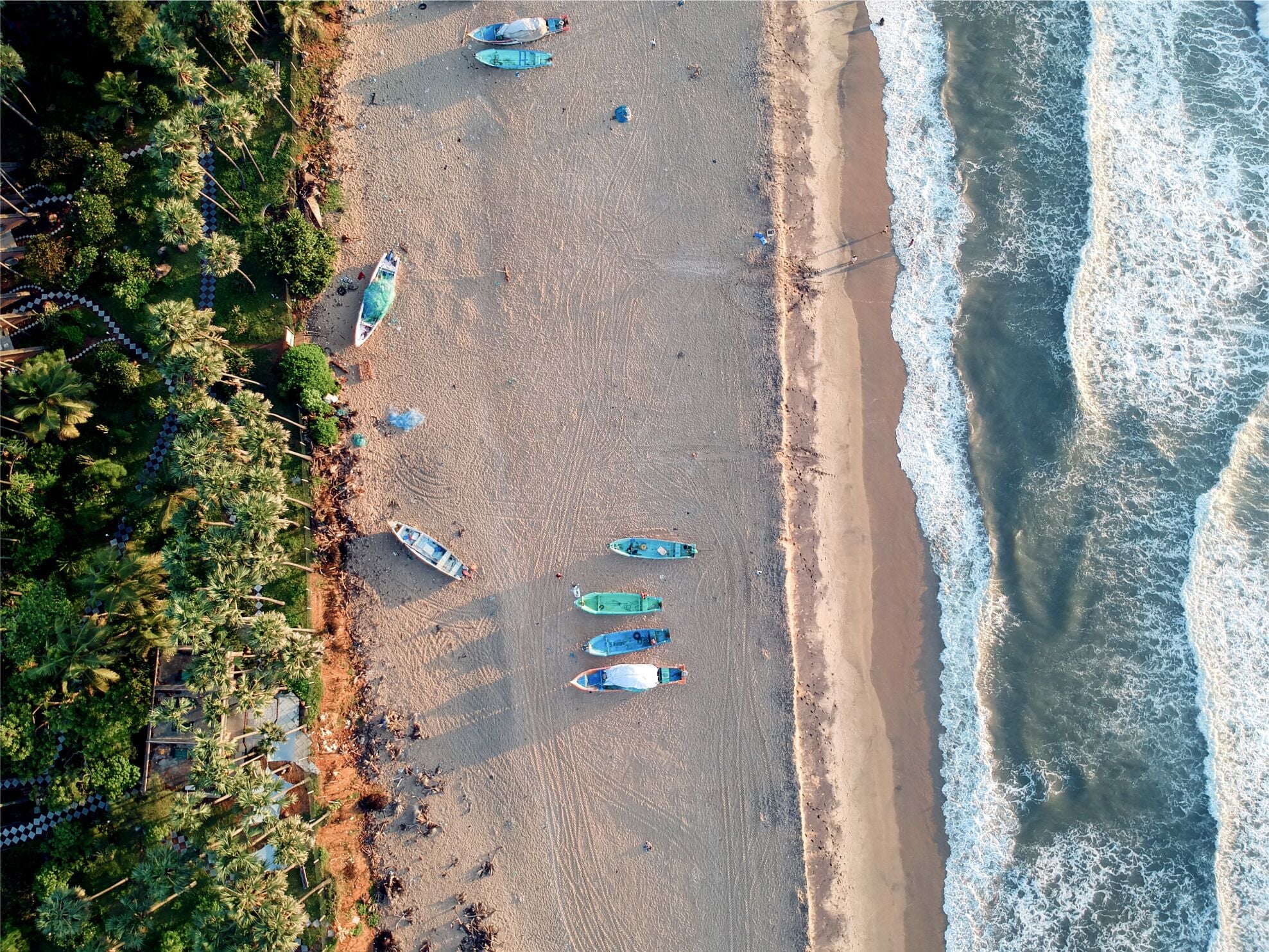 Vista aérea de uma praia com vários barcos enfileirados na areia, cercados por vegetação densa.