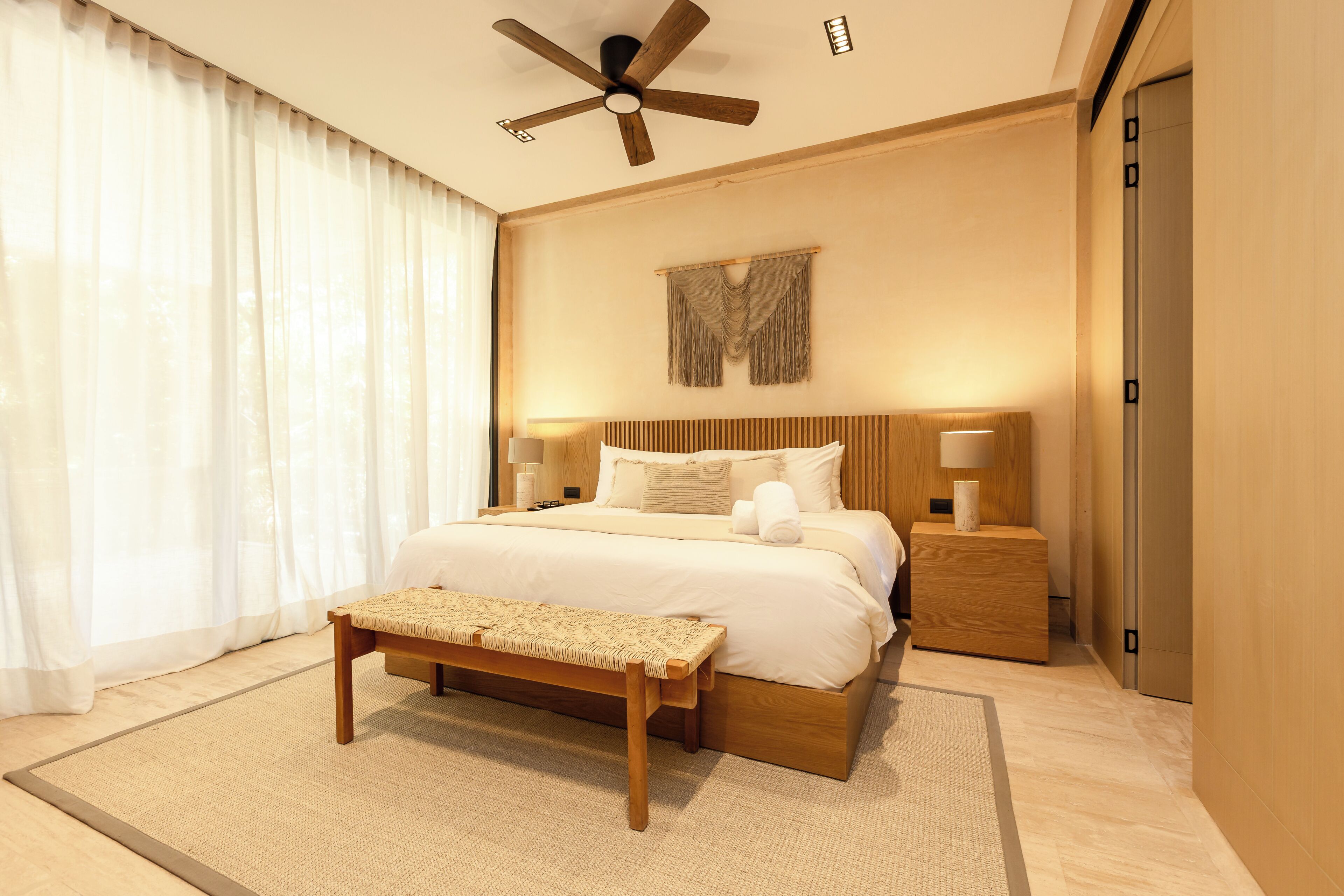 Quarto de hotel moderno com cama grande, cabeceira de madeira, luminárias laterais, um banco e ventilador de teto.
