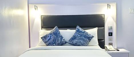 Uma cama bem arrumada com um cobre-leito estampado e duas almofadas, uma mesinha de cabeceira com um telefone e uma luminária fixada na parede.