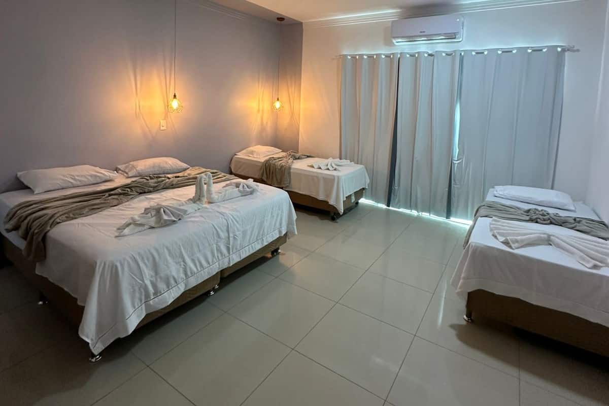 Quarto de hotel com duas camas, uma janela grande com cortinas, um aparelho de ar condicionado e luminárias fixadas na parede.