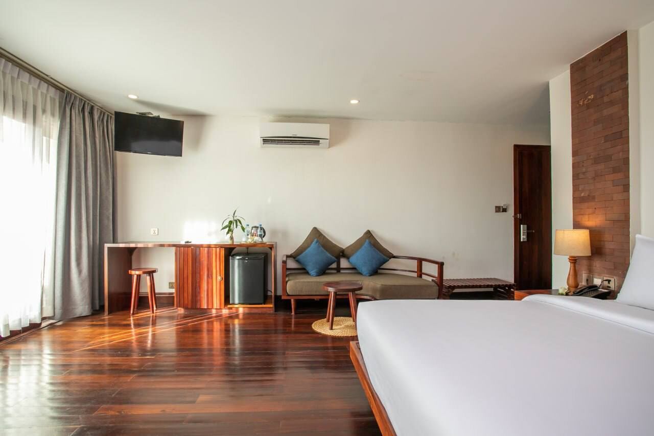 Quarto de hotel com cama, escrivaninha com cadeira, pequena cozinha equipada, sofá com almofadas e televisão na parede.