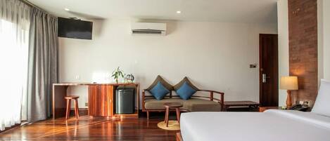 Quarto de hotel com cama, escrivaninha com cadeira, pequena cozinha equipada, sofá com almofadas e televisão na parede.