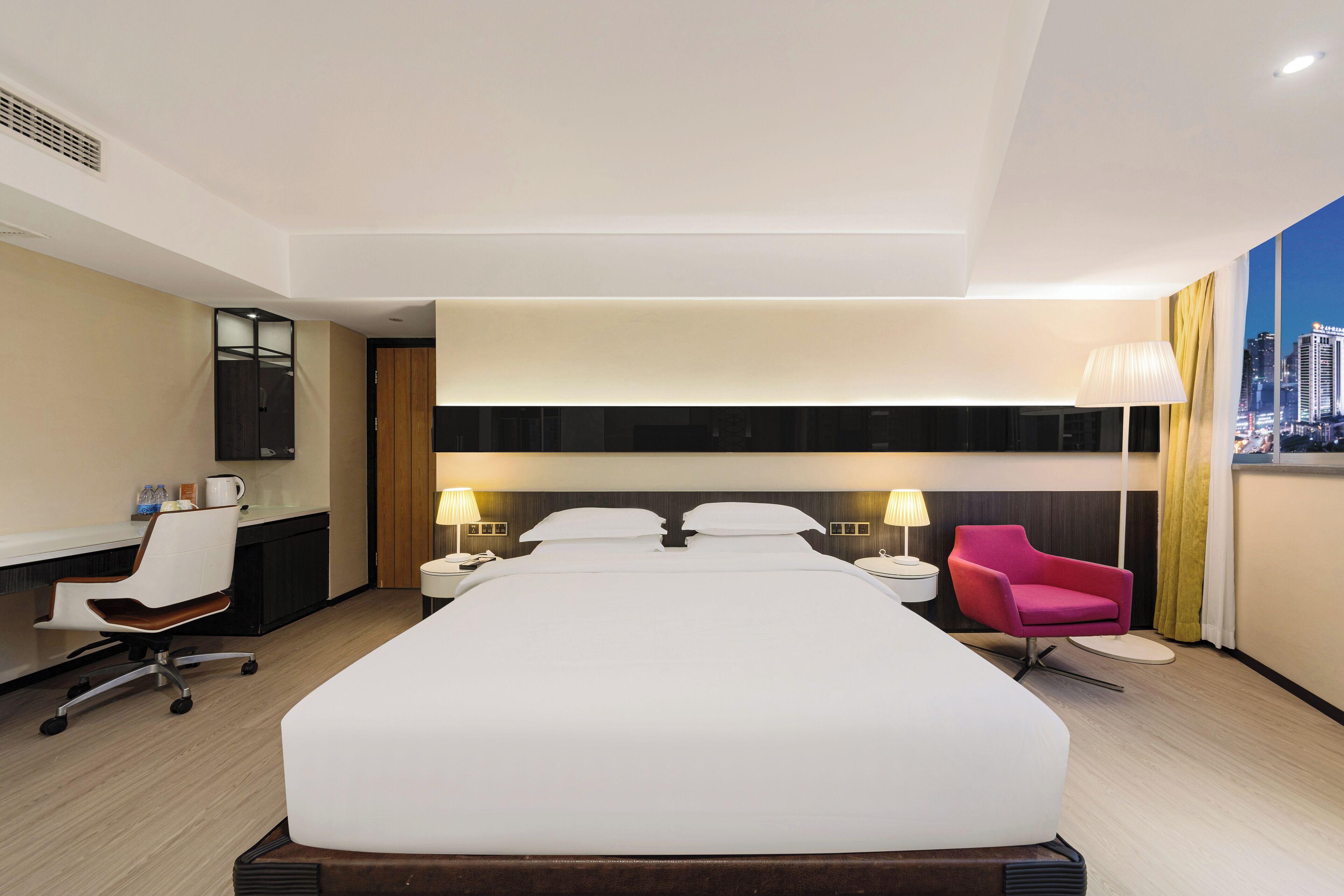 Quarto de hotel moderno com cama grande, escrivaninha, cadeira e vista para a cidade.