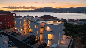 Um edifício moderno com várias varandas, área de estacionamento e vista para a água e montanhas ao entardecer.