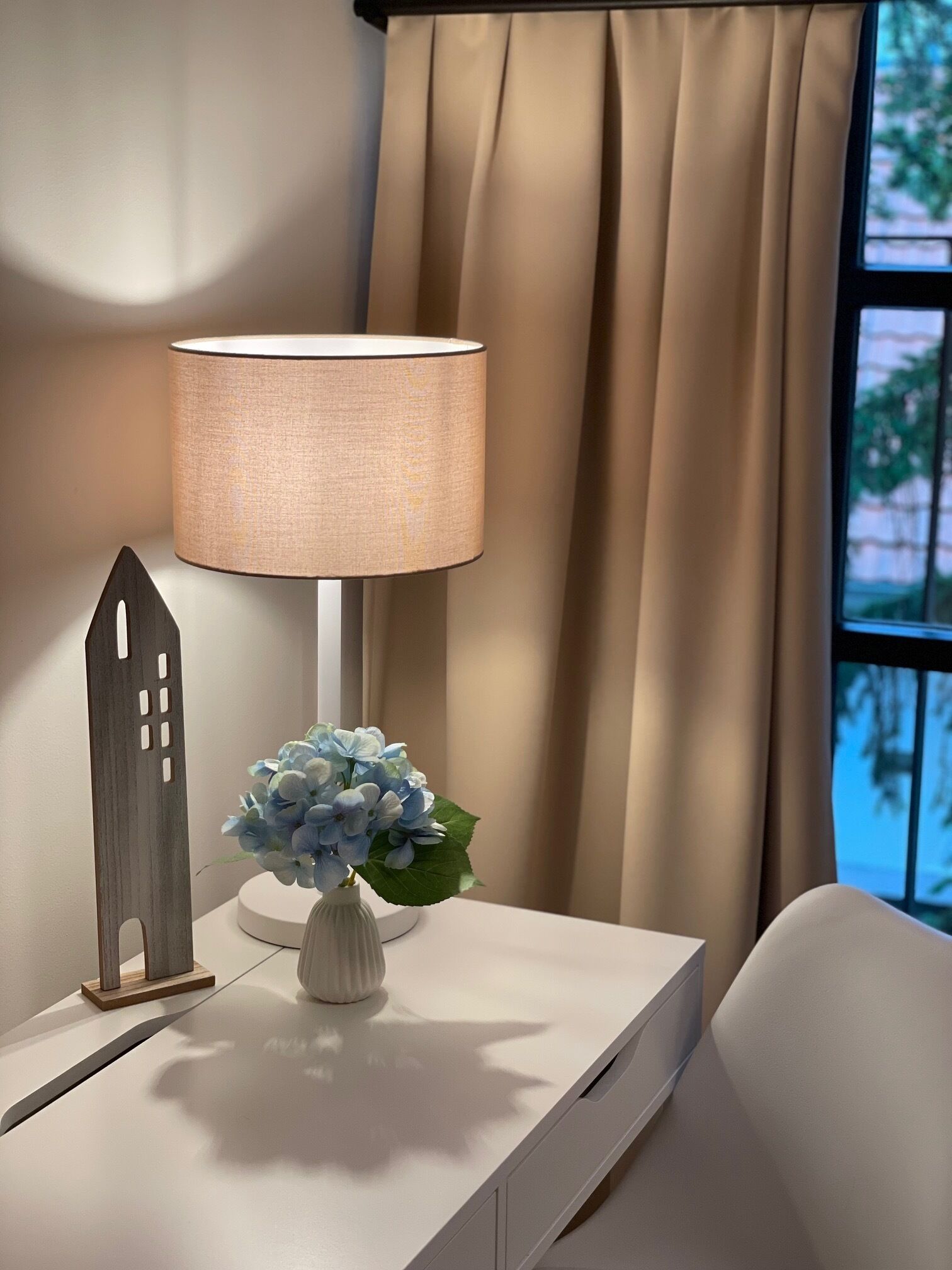 Quarto moderno com mesa de cabeceira branca, luminária com cúpula bege, objeto de madeira decorativo e um vaso com flores azuis.