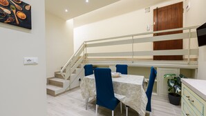 Uma sala de jantar com uma mesa posta para quatro pessoas, cadeiras azuis, uma escada e uma porta de madeira ao fundo.