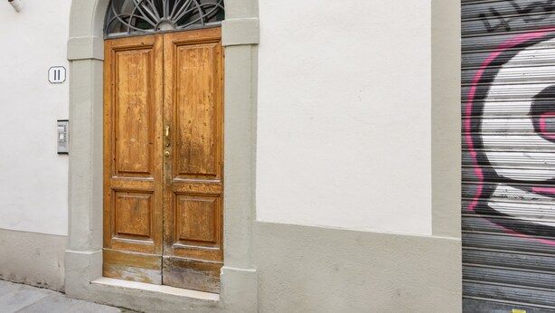Uma porta dupla de madeira com uma pequena janela acima, em um edifício branco com uma persiana de metal à direita.