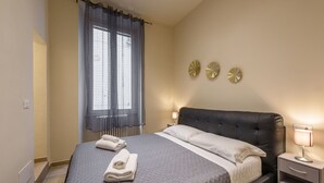 Cama bem arrumada com colcha cinza texturizada, dois travesseiros e toalhas dobradas ao lado. Há uma mesa de cabeceira com uma luminária, e três placas decorativas na parede acima da cama.