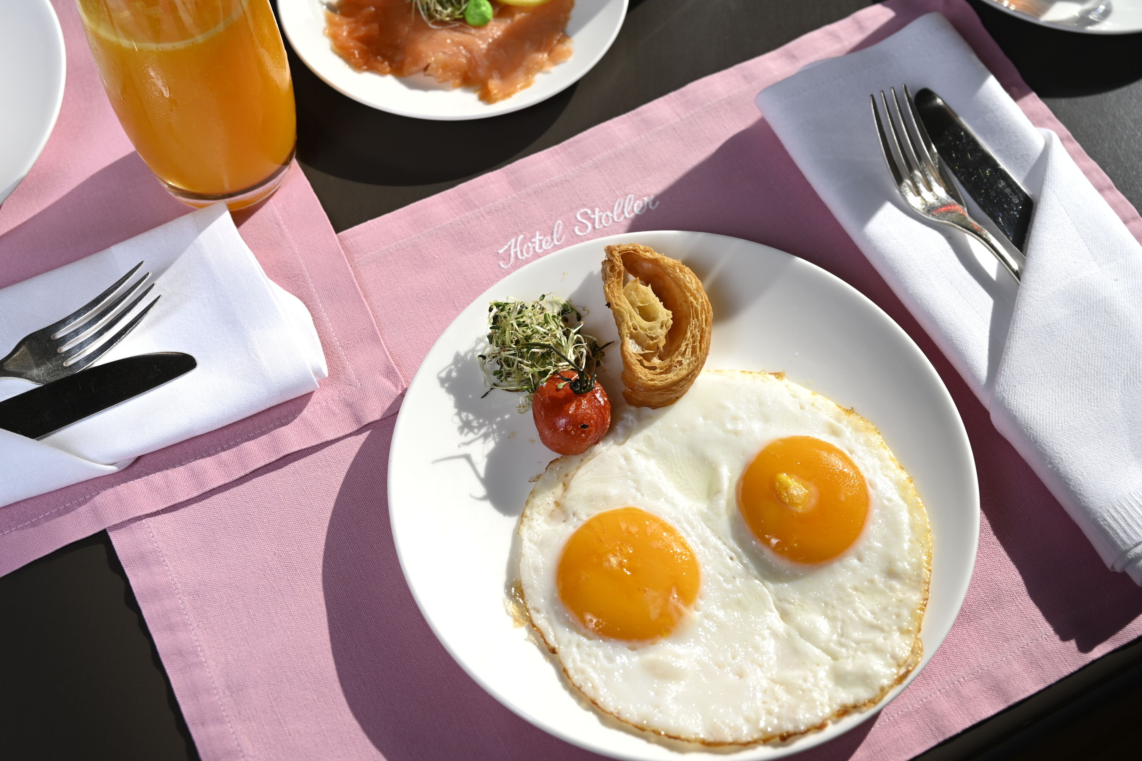 Prato de café da manhã com dois ovos fritos, um tomate pequeno e um doce, acompanhados por garfo e faca sobre uma toalha de mesa rosa, com um copo de suco de laranja ao fundo.