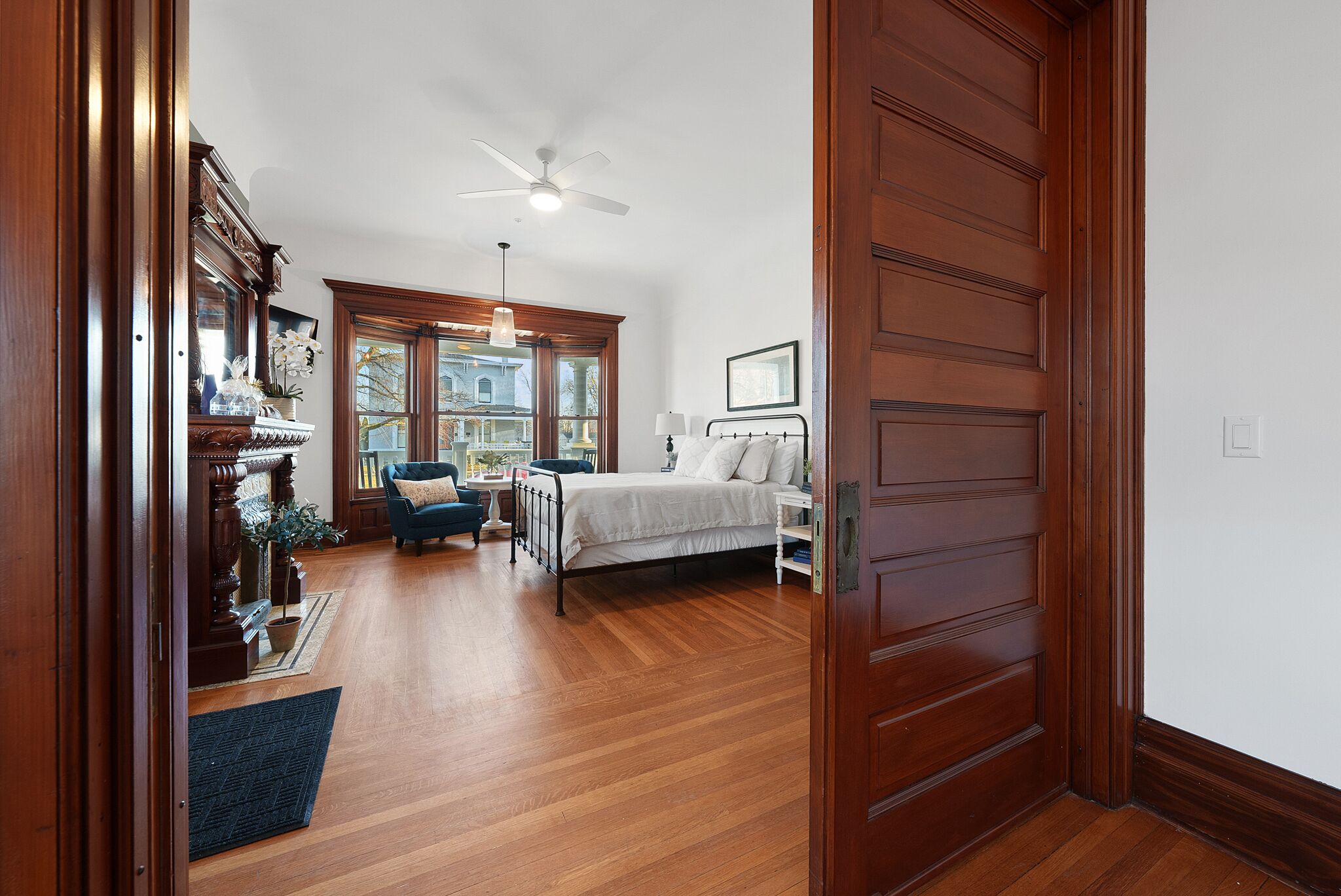 Quarto com piso de madeira, cama, mesa de cabeceira, cômoda, ventilador de teto e janela com vista para um edifício.