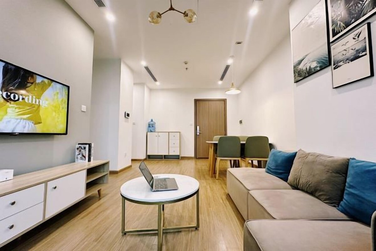 Uma sala de estar moderna com um sofá, uma mesa de centro redonda com um laptop, uma TV na parede e uma área de jantar com cadeiras e uma mesa.