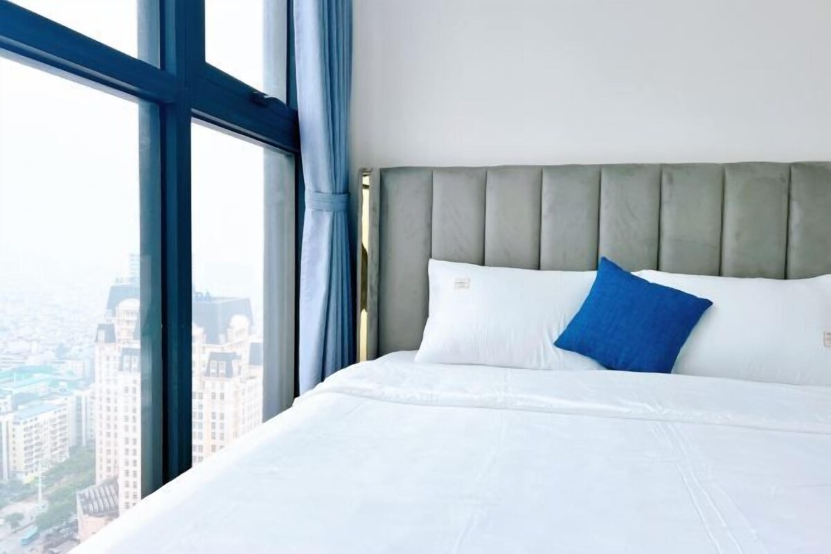 Quarto de hotel com uma janela grande, cabeceira cinza estofada, colcha branca e duas almofadas (uma branca, uma azul).