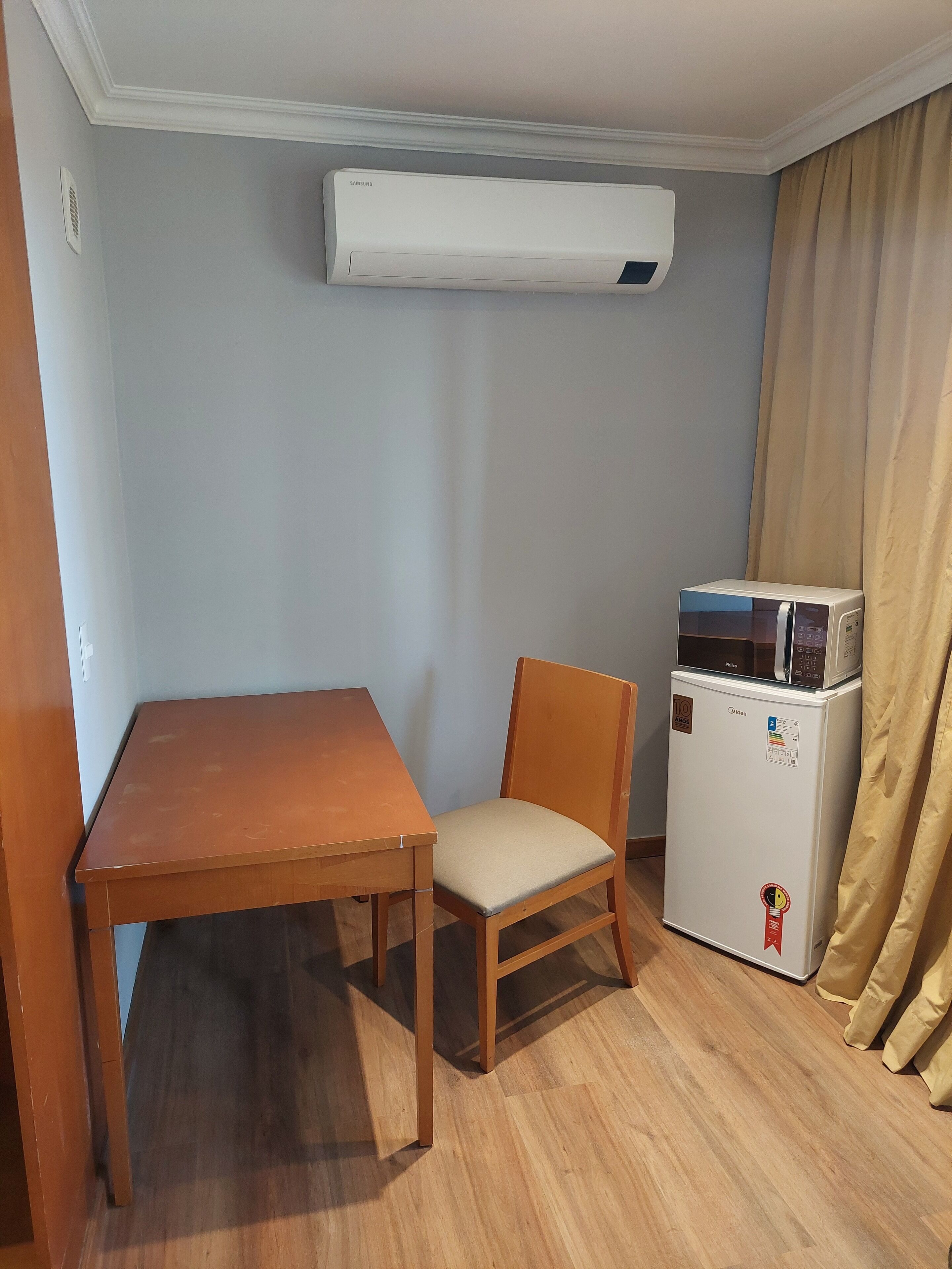 Um quarto pequeno com uma mesa e cadeira de madeira, uma geladeira branca com micro-ondas e um aparelho de ar condicionado na parede.