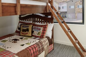 Uma cama beliche com colcha xadrez e duas almofadas com estampas de ursos. Uma escada de madeira está encostada à parede.