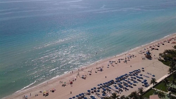 Uma praia com fileiras de guarda-sóis e cadeãsas de praia, margem arenosa e o oceano ao fundo.