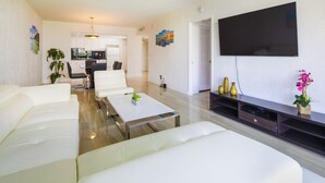 Sala de estar moderna com sofás brancos, televisão de tela plana e cozinha ao fundo.