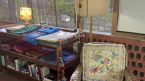 Um quarto com uma cadeira de fibras naturais, uma mesa com cobertores dobrados e uma estante repleta de livros.
