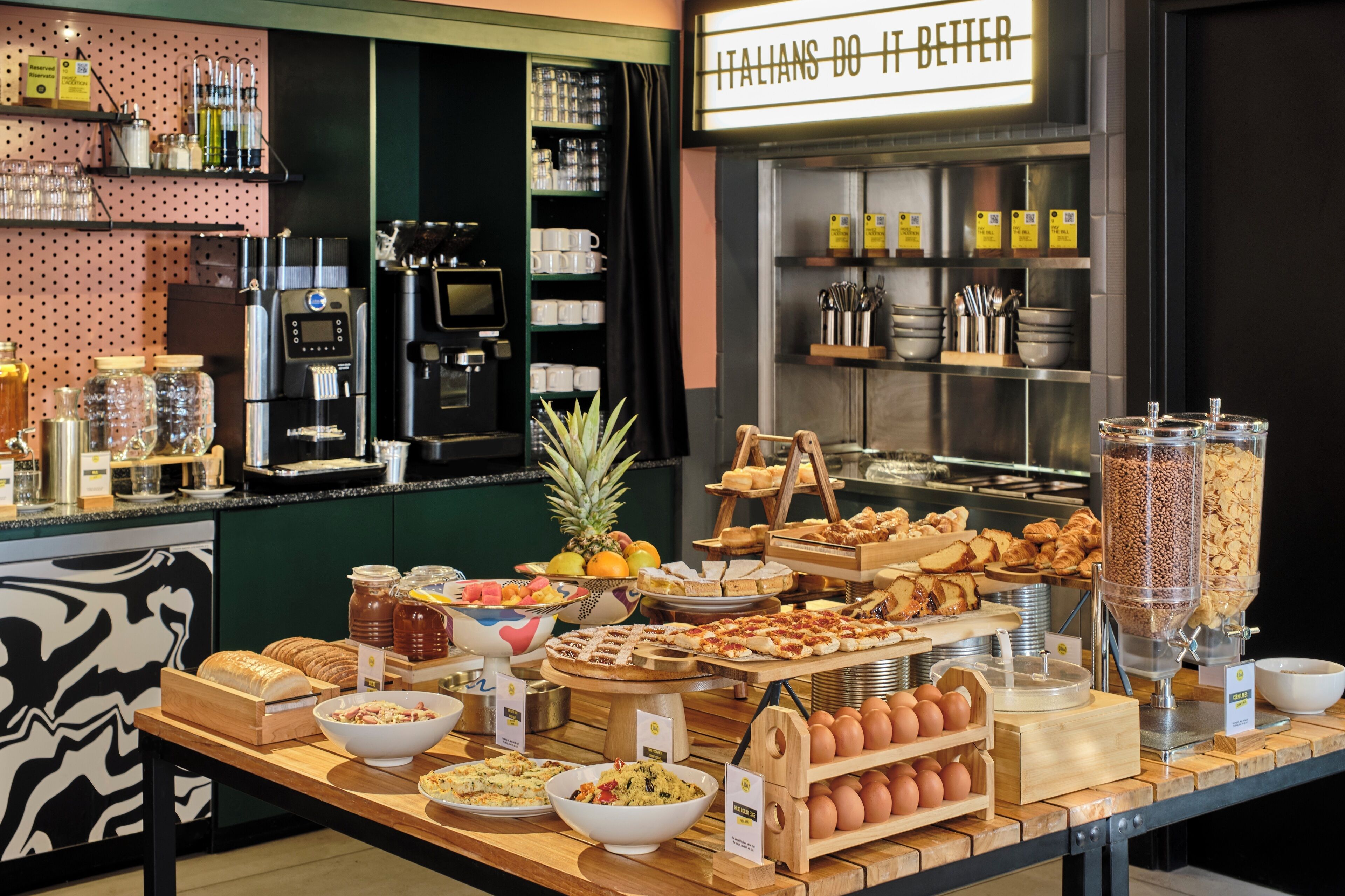 Buffet de café da manhã com diversos alimentos, máquinas de café e uma placa com a inscrição 'Italians do it better'.