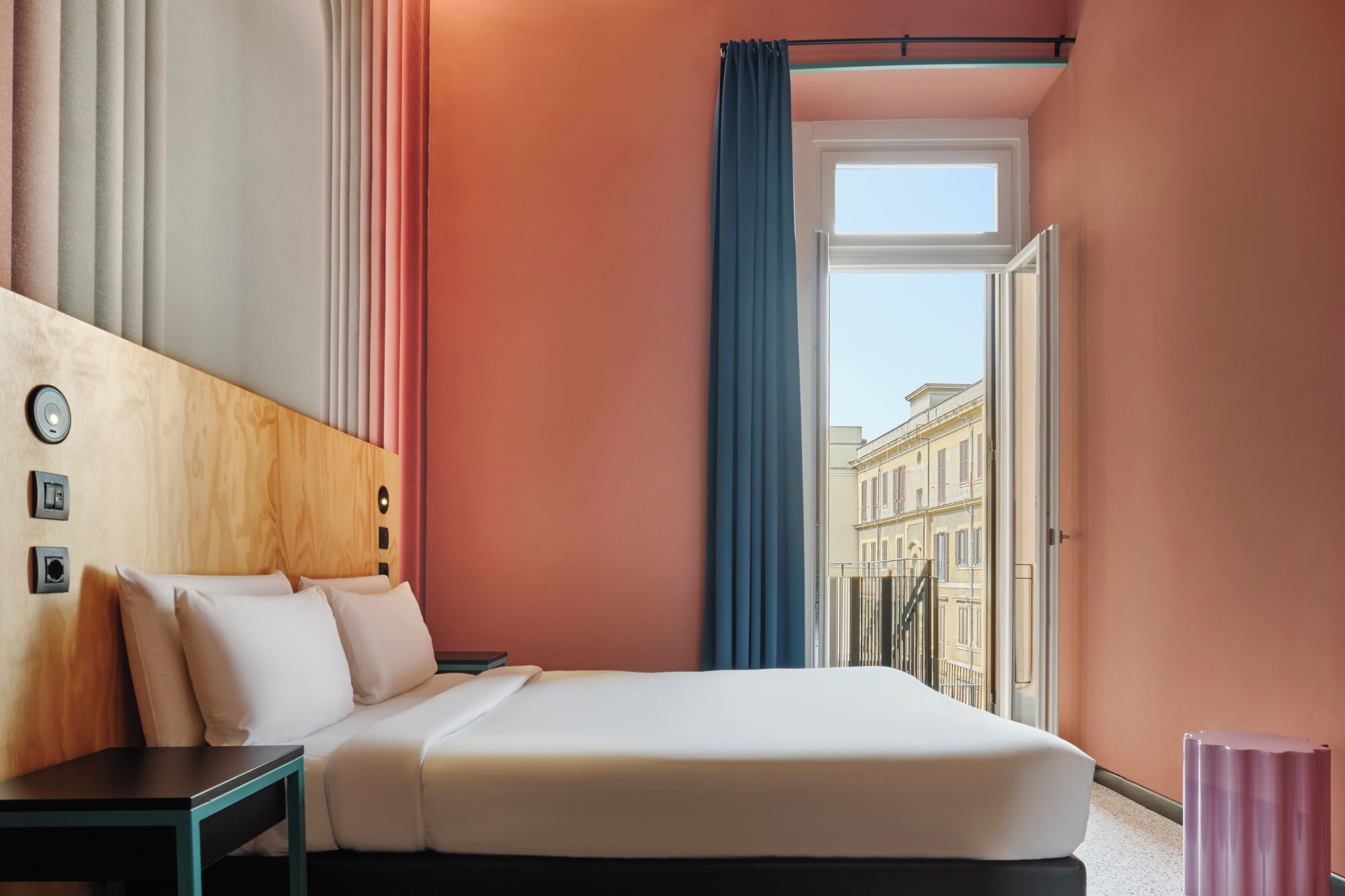 Quarto de hotel com cama, cabeceira de madeira, mesa de cabeceira, janela com cortinas e vista para prédios.