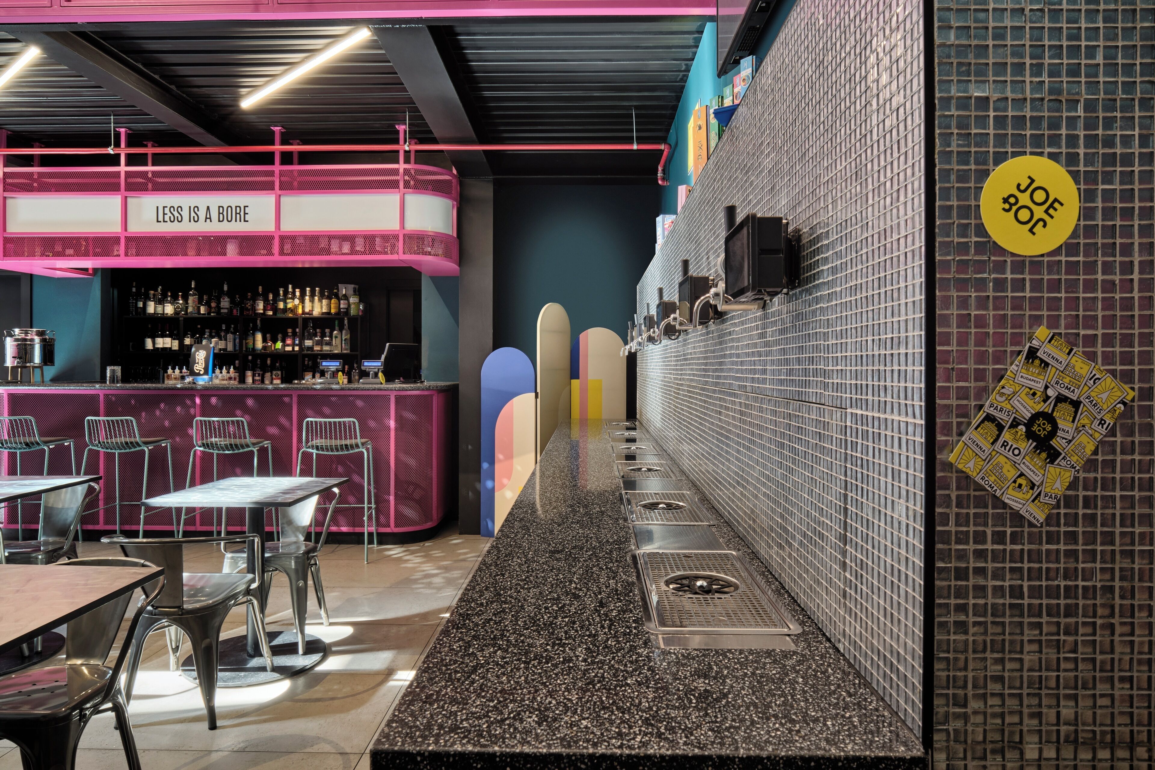 Uma área de bar moderna com balcão rosa, cadeiras de metal e uma parede revestida com um letreiro 'JOE BOC'.