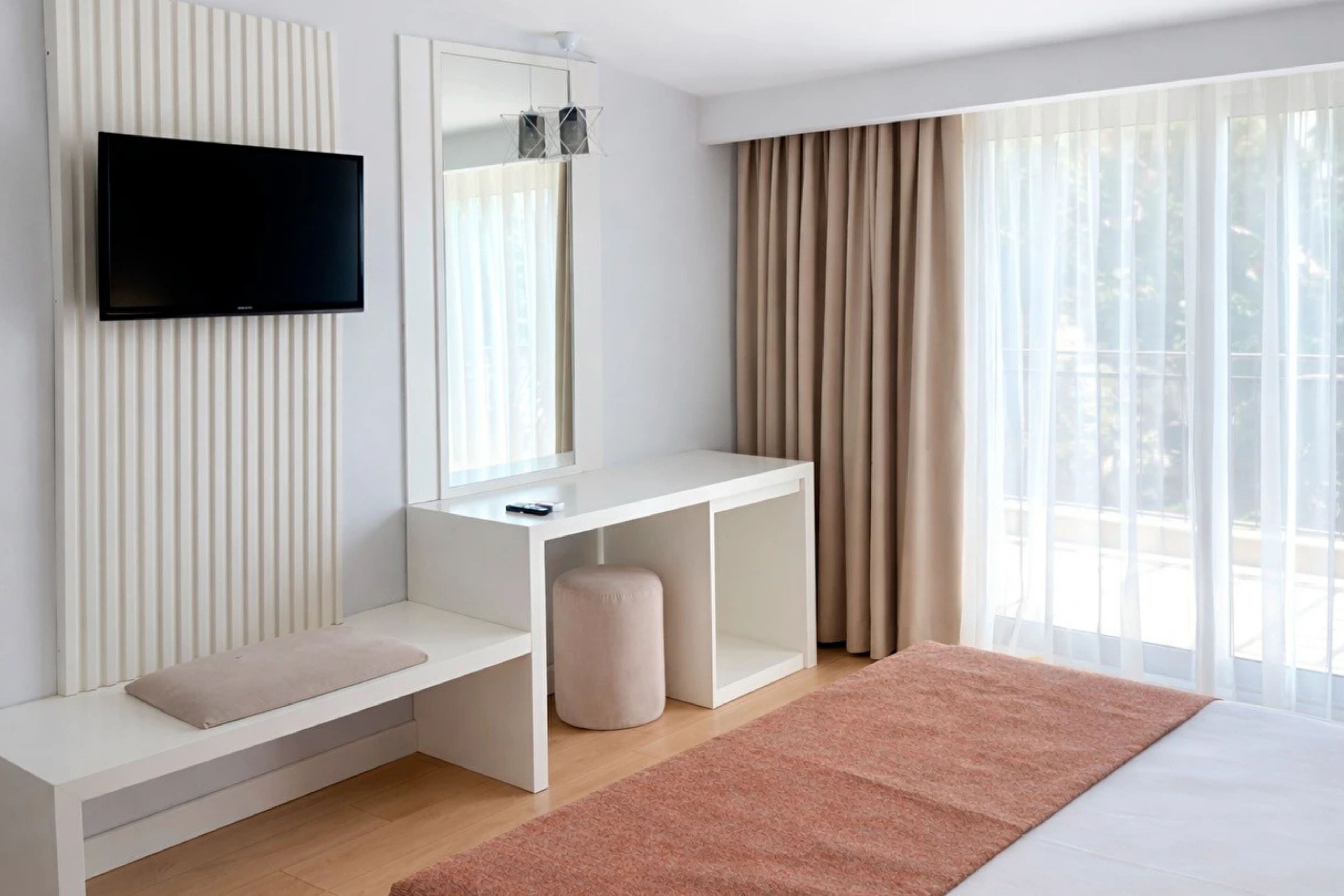 Quarto de hotel moderno com escrivaninha branca, televisão de tela plana, janela grande com cortinas e uma cama espaçosa.