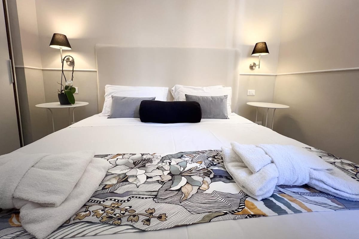 Quarto de hotel com uma cama grande, duas mesas de cabeceira e duas luminárias fixadas na parede.