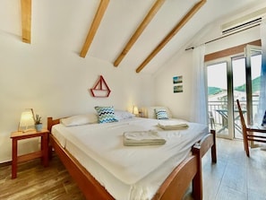 Quarto com cama de madeira, mesa de cabeceira, decoração geométrica vermelha na parede e vista para uma varanda com móveis de exterior.