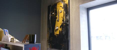 Quarto com uma grande pintura abstrata na parede, uma escrivaninha com computador e uma janela.
