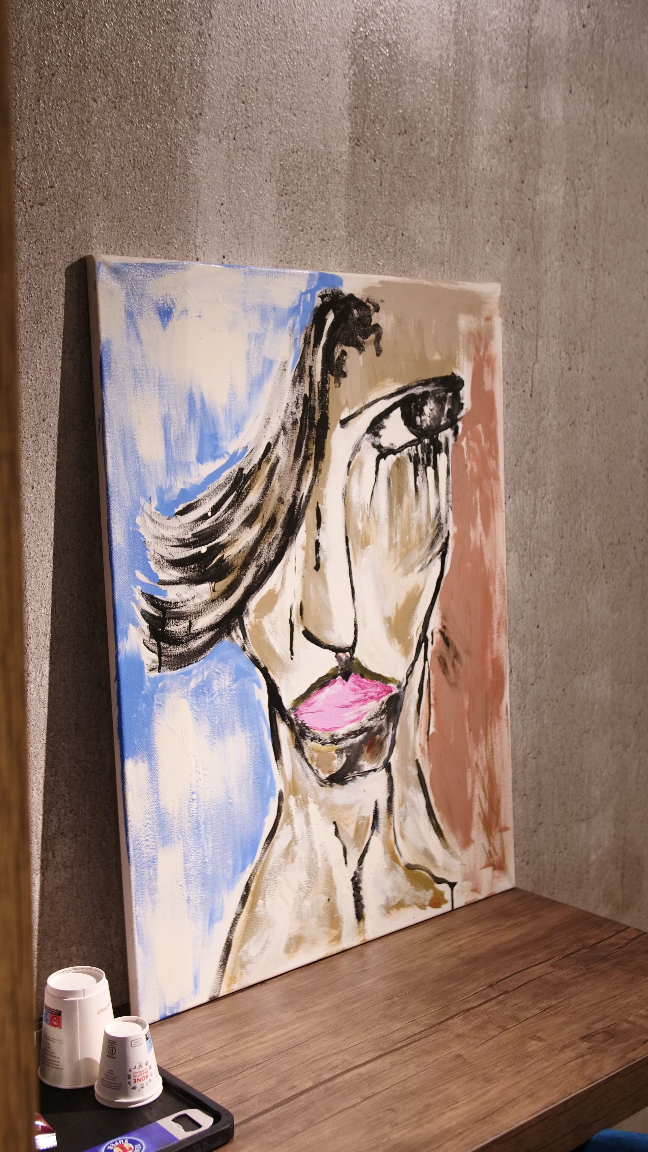 Pintura do rosto de uma mulher em tela, apoiada sobre uma mesa de madeira em frente a uma parede texturizada.