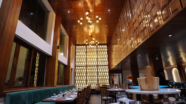 Restaurante com painéis de madeira, teto alto e luminária central. Mesas montadas com talheres e copos.