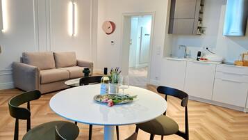 Uma sala de estar moderna com uma mesa redonda, duas cadeiras, um sofá e uma área de cozinha com geladeira e armários.