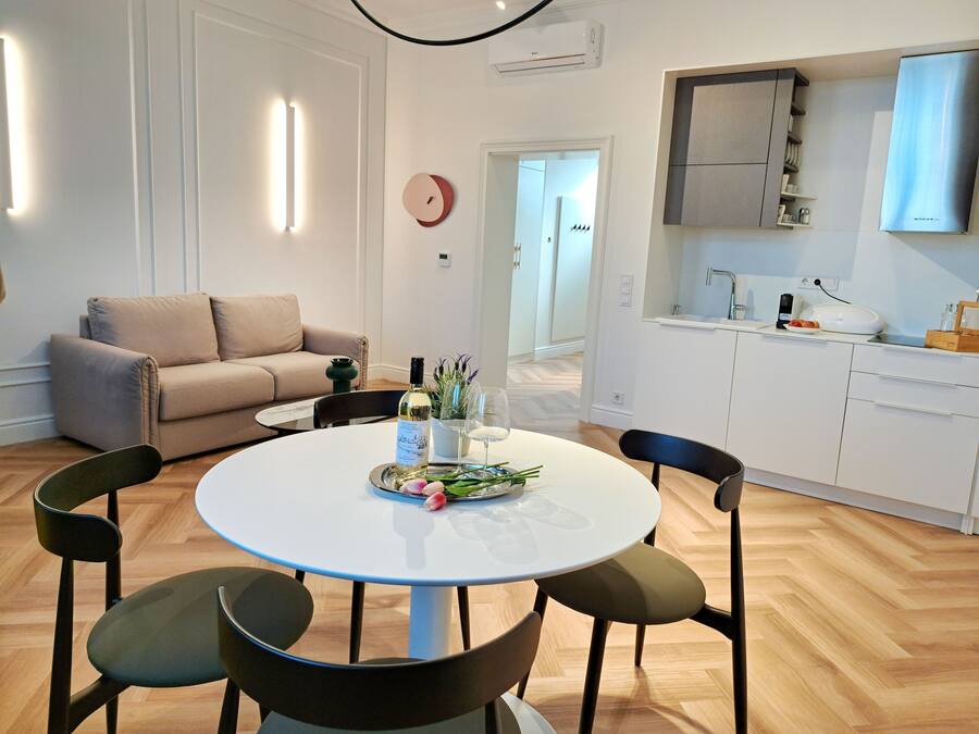 Uma sala de estar moderna com uma mesa redonda, duas cadeiras, um sofá e uma área de cozinha com geladeira e armários.