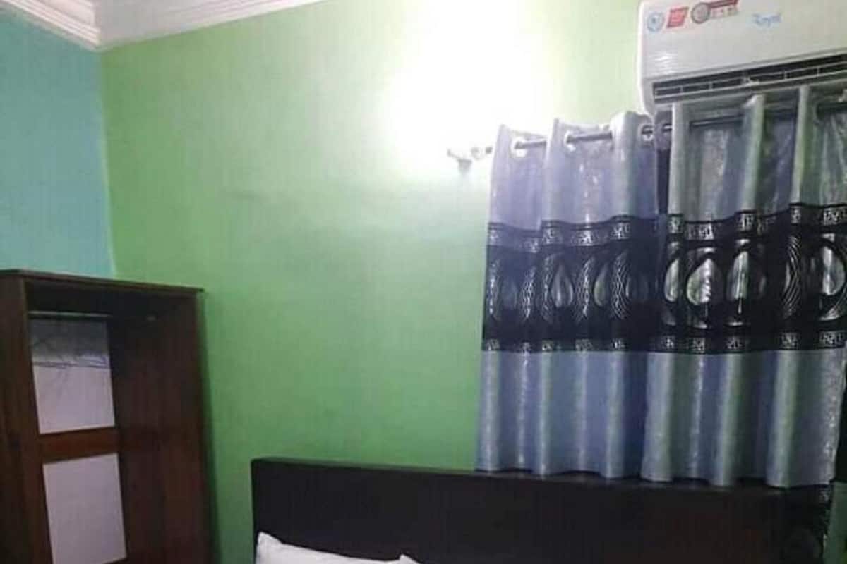 Uma cama com almofadas brancas e um cobertor azul e vermelho, um guarda-roupa de madeira e um ar condicionado fixado na parede.