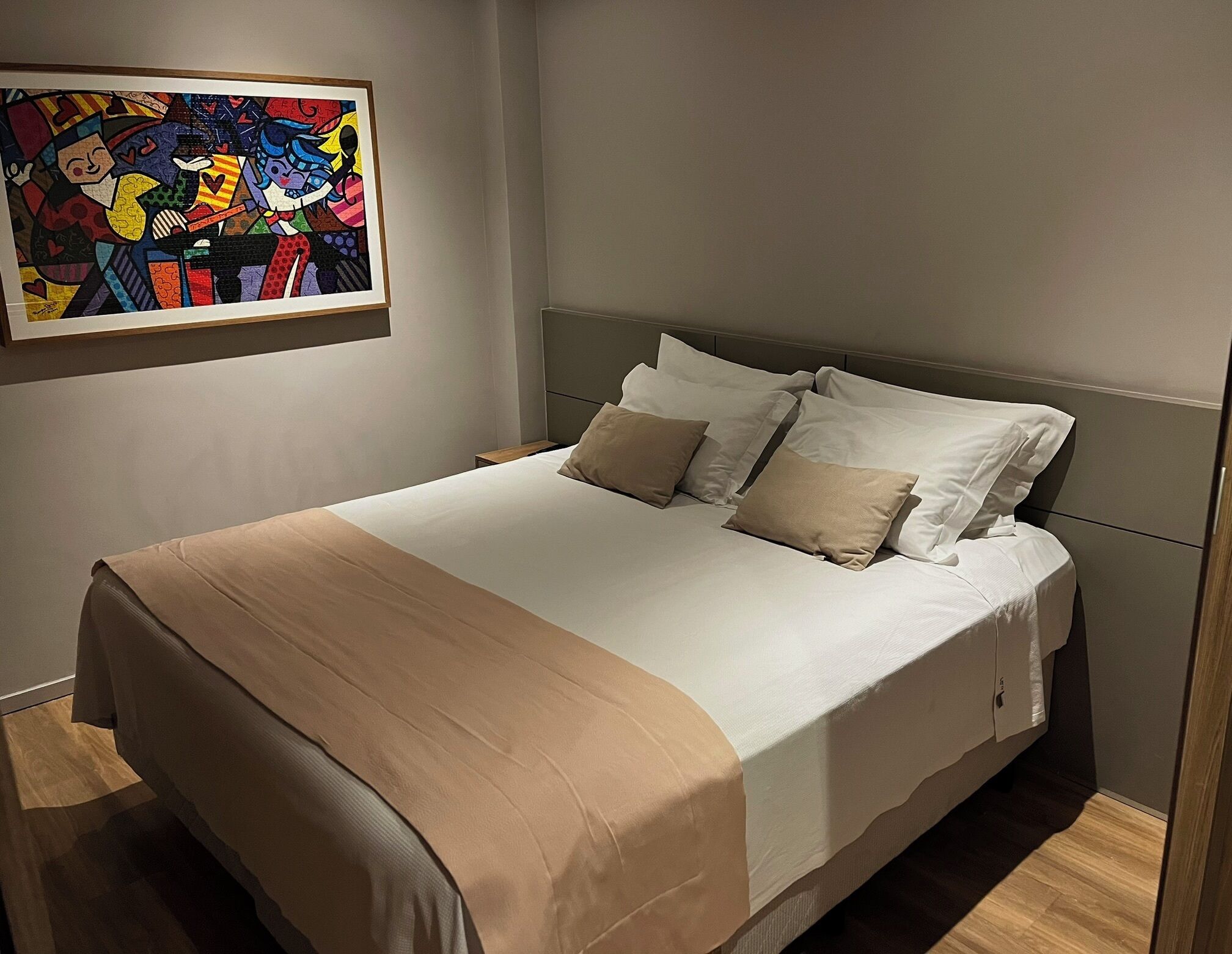Cama bem arrumada com colcha bege e travesseiros, cabeceira e mesa de cabeceira pequena com abajur.