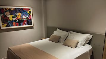 Cama bem arrumada com colcha bege e travesseiros, cabeceira e mesa de cabeceira pequena com abajur.